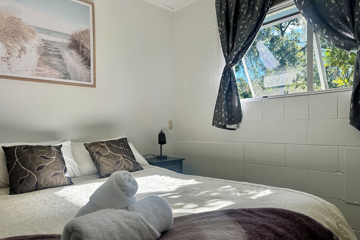 Cama bem arrumada com manta branca e marrom, duas almofadas e toalha branca enrolada. Um quadro com cena de praia na parede, e janela com cortinas visível.