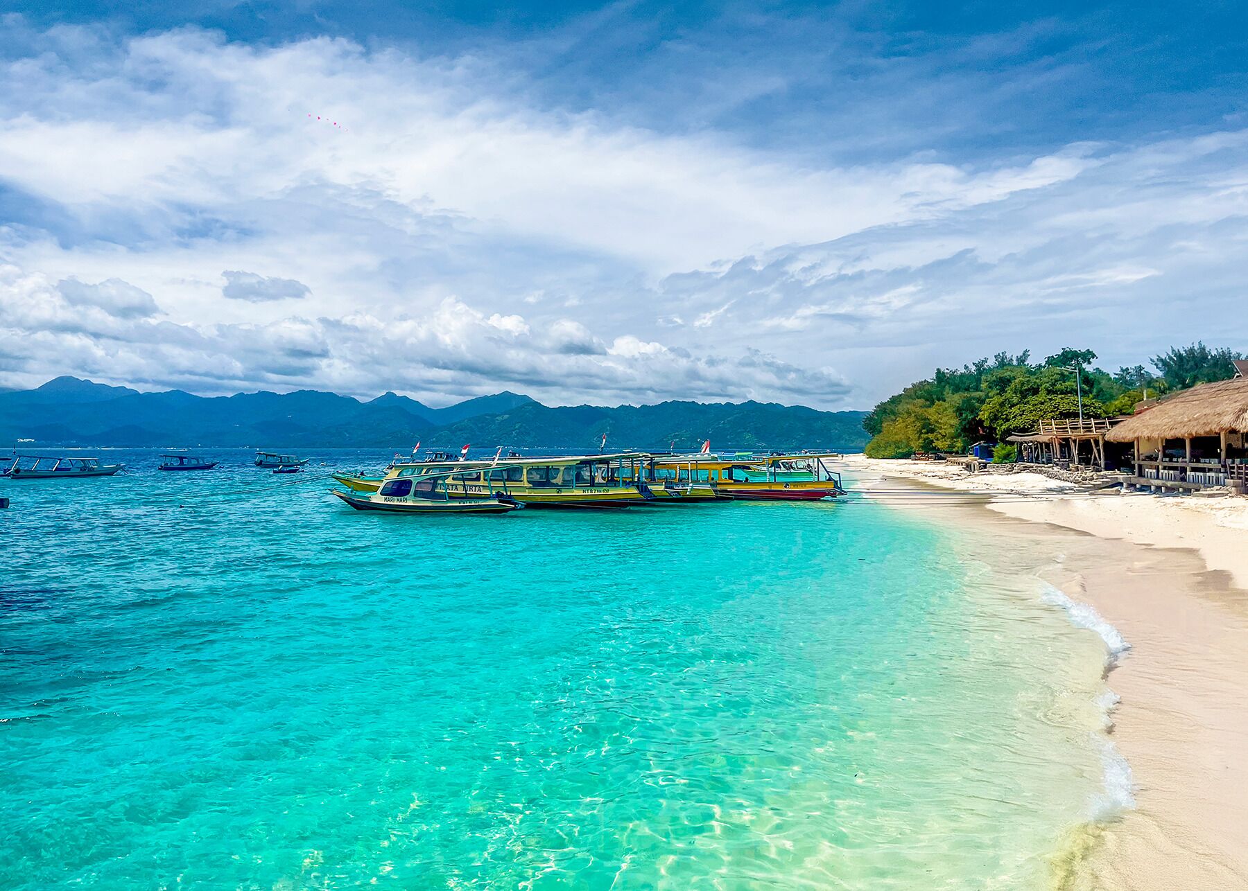 Uma praia com águas turquesa cristalinas, um barco atracado perto da costa e montanhas ao fundo.