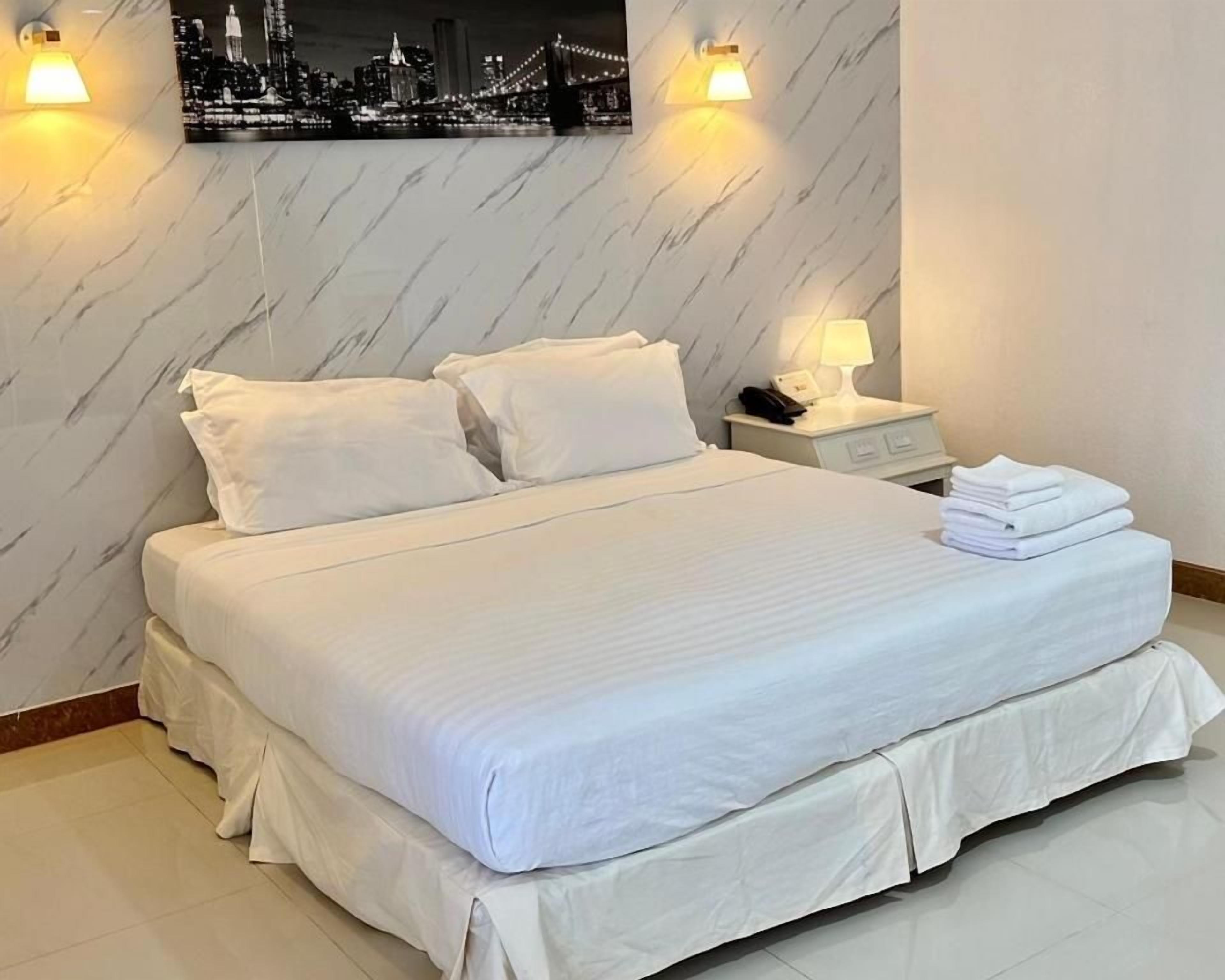 Cama bem arrumada com lençóis brancos em quarto de hotel com paredes em estilo marmorizado e duas luminárias.