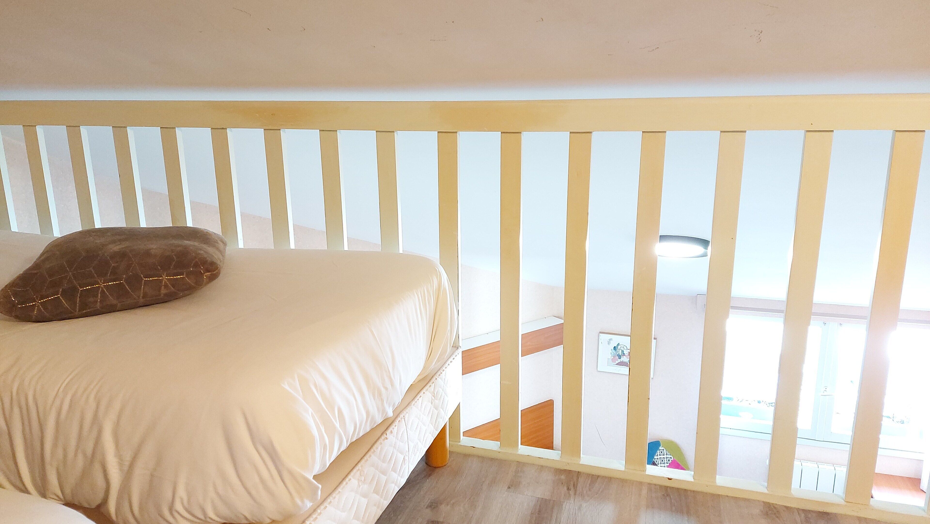 Quarto com cama, travesseiro, corrimão de madeira e vista para o exterior.