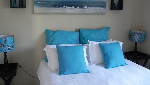 Quarto com cama, almofadas azuis e brancas, pintura do oceano e duas mesas de cabeceira com abajures.