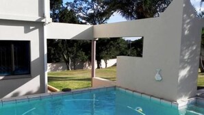 Uma piscina com design arquitetônico moderno, cercada por um edifício branco e uma área gramada com árvores.
