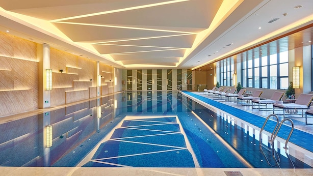 Piscina interior moderna com iluminação de teto geométrica, espreguiçadeiras e vista clara da piscina.