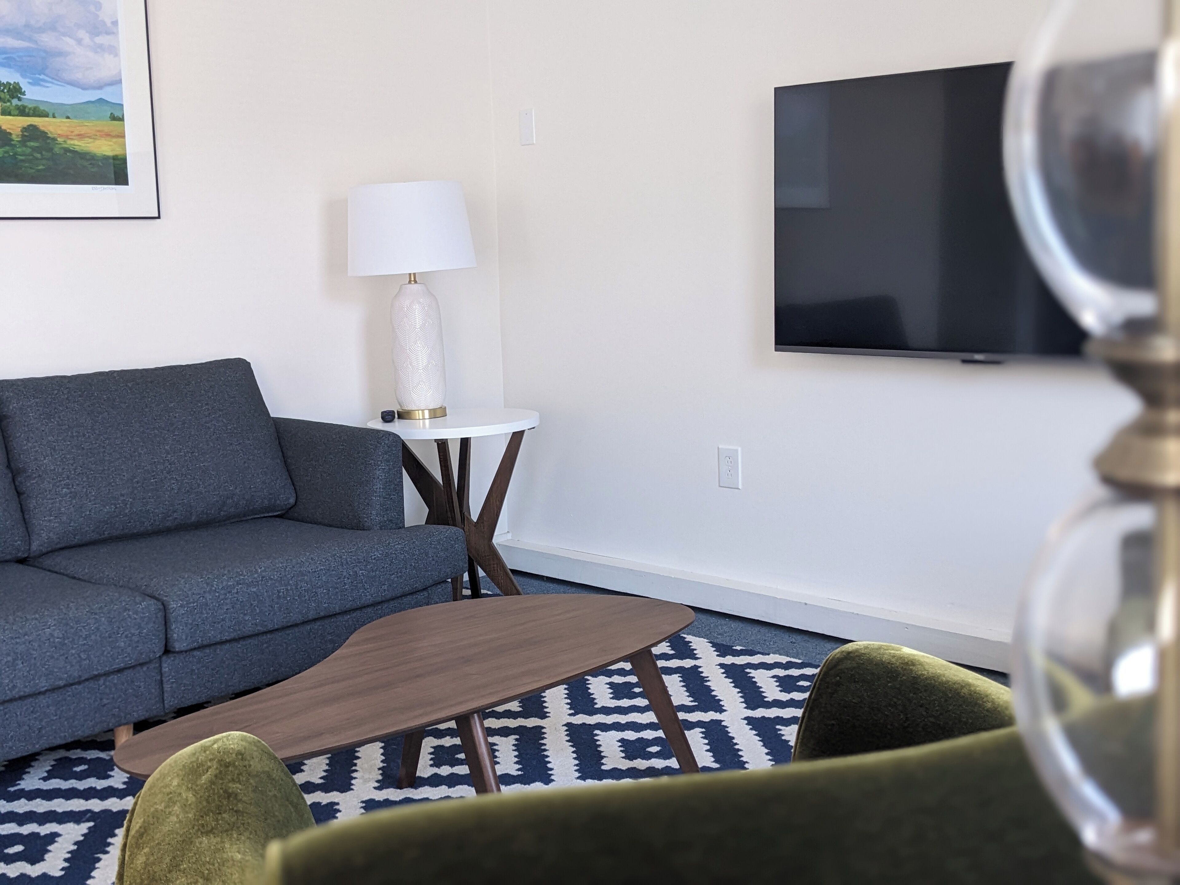 Uma sala de estar moderna com um sofá cinzento, uma mesinha branca com uma luminária, um tapete estampado e uma TV na parede.