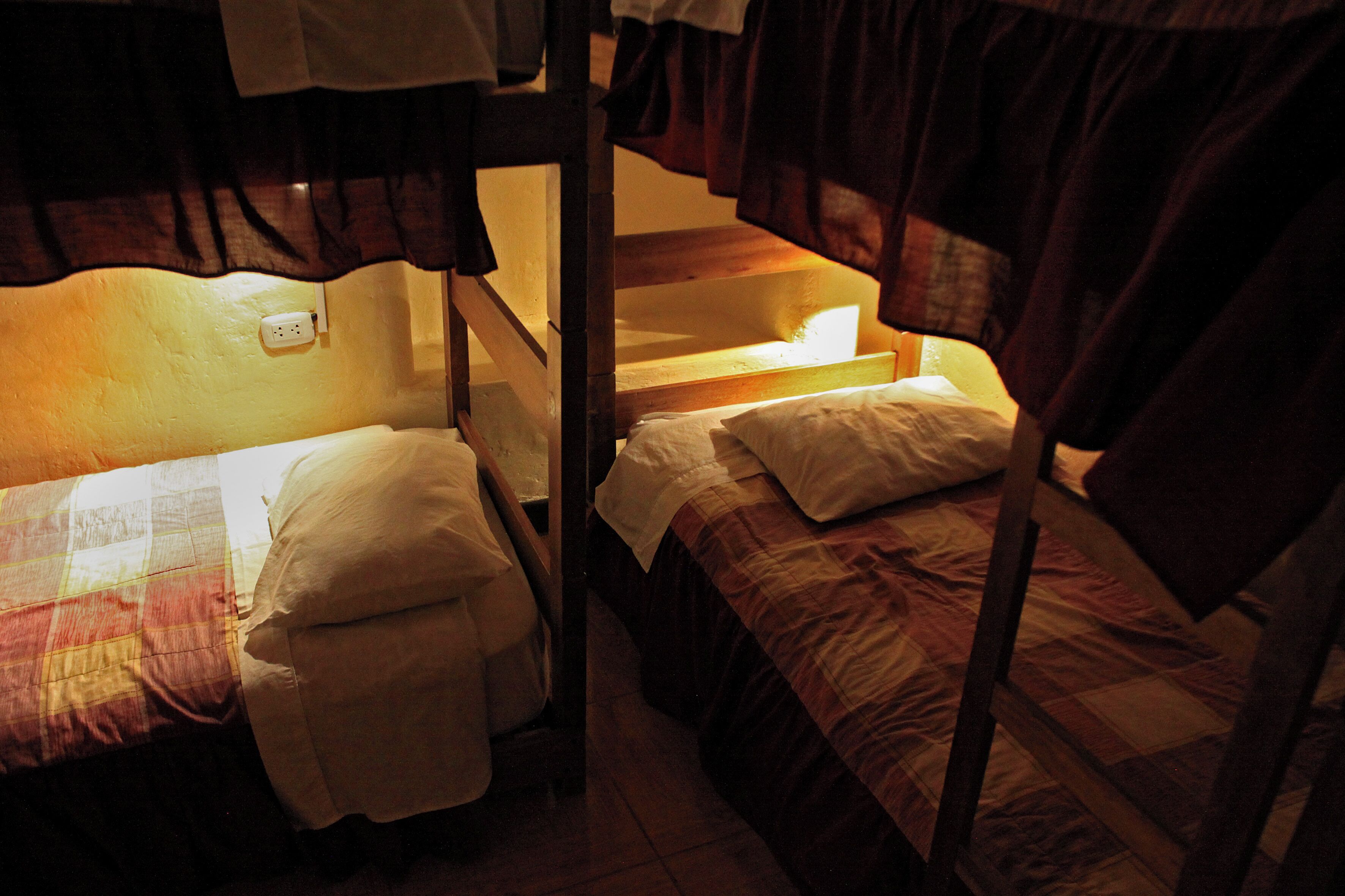 Quarto de dormitório com beliches, uma tomada visível e uma janela com cortinas.