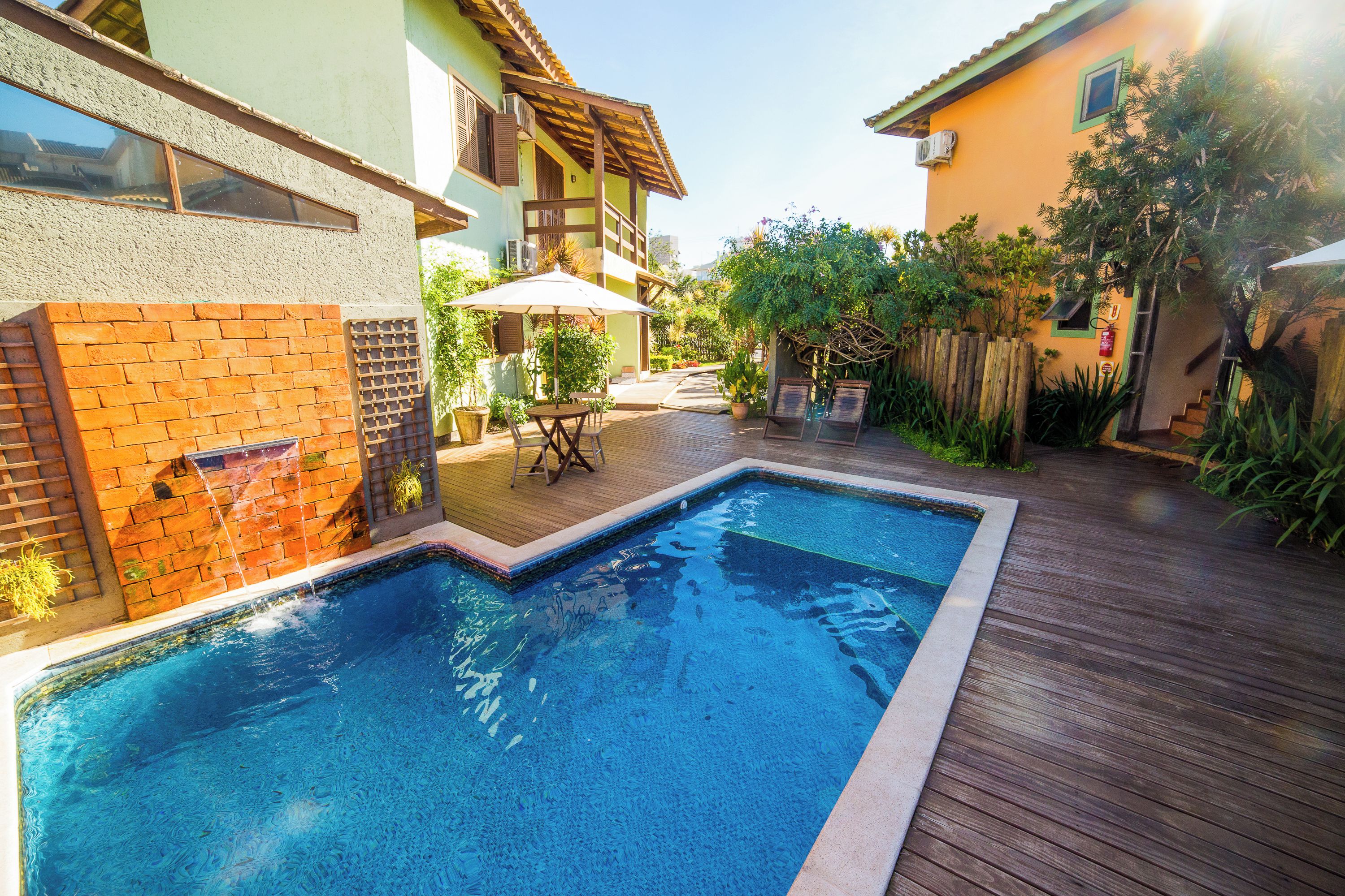 Piscina com deck de madeira, cercada por edifícios e vegetação.