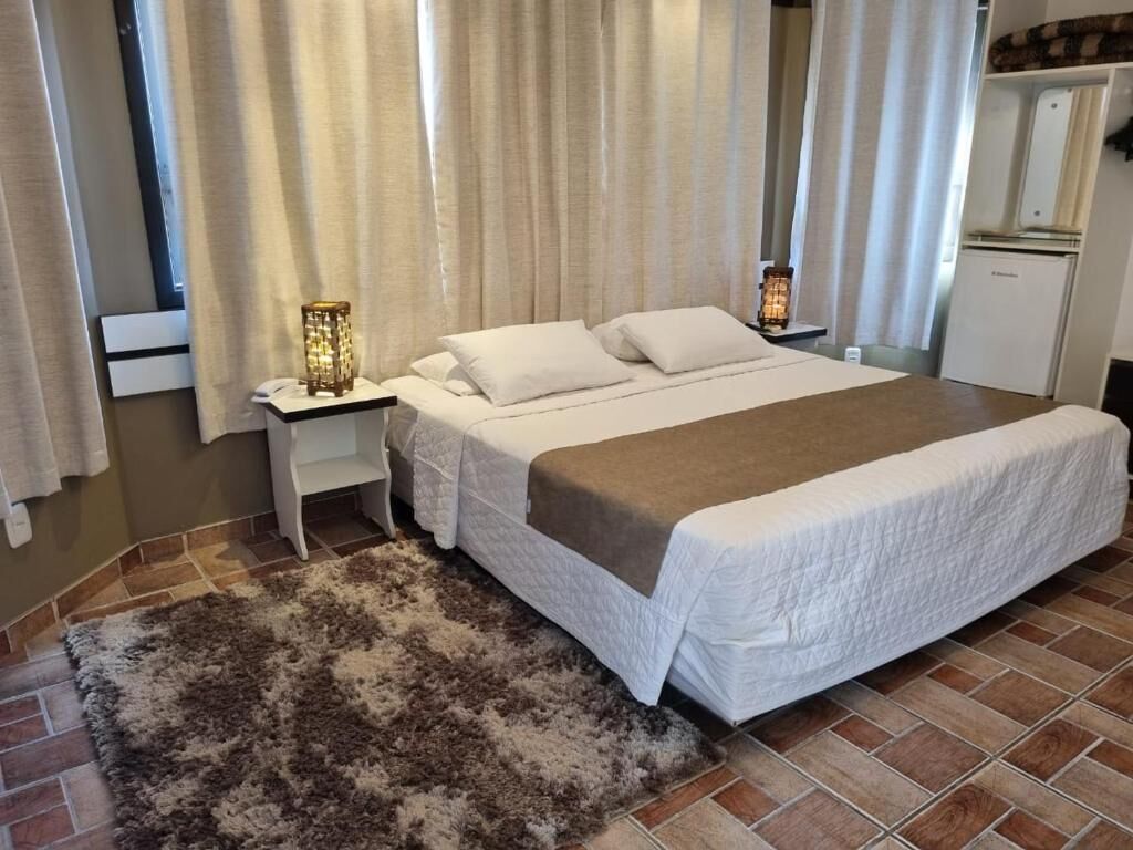 Quarto de hotel com cama, mesas de cabeceira, luminária, geladeira e tapete.