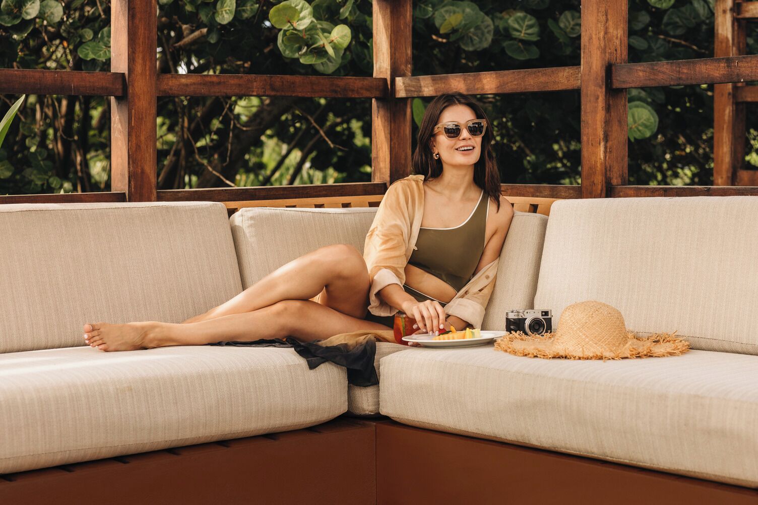 Uma pessoa sentada em um sofá bege, com uma câmera e um chapéu de palha no chão ao lado.