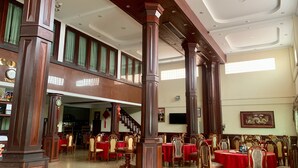 Um salão amplo com altas colunas de madeira, várias mesas de jantar com toalhas vermelhas e uma escadaria que leva a um andar superior.