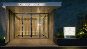 Entrada do Hotel Sakura Suite, com fachada de vidro moderna e uma placa exibindo o nome e a localização do hotel.