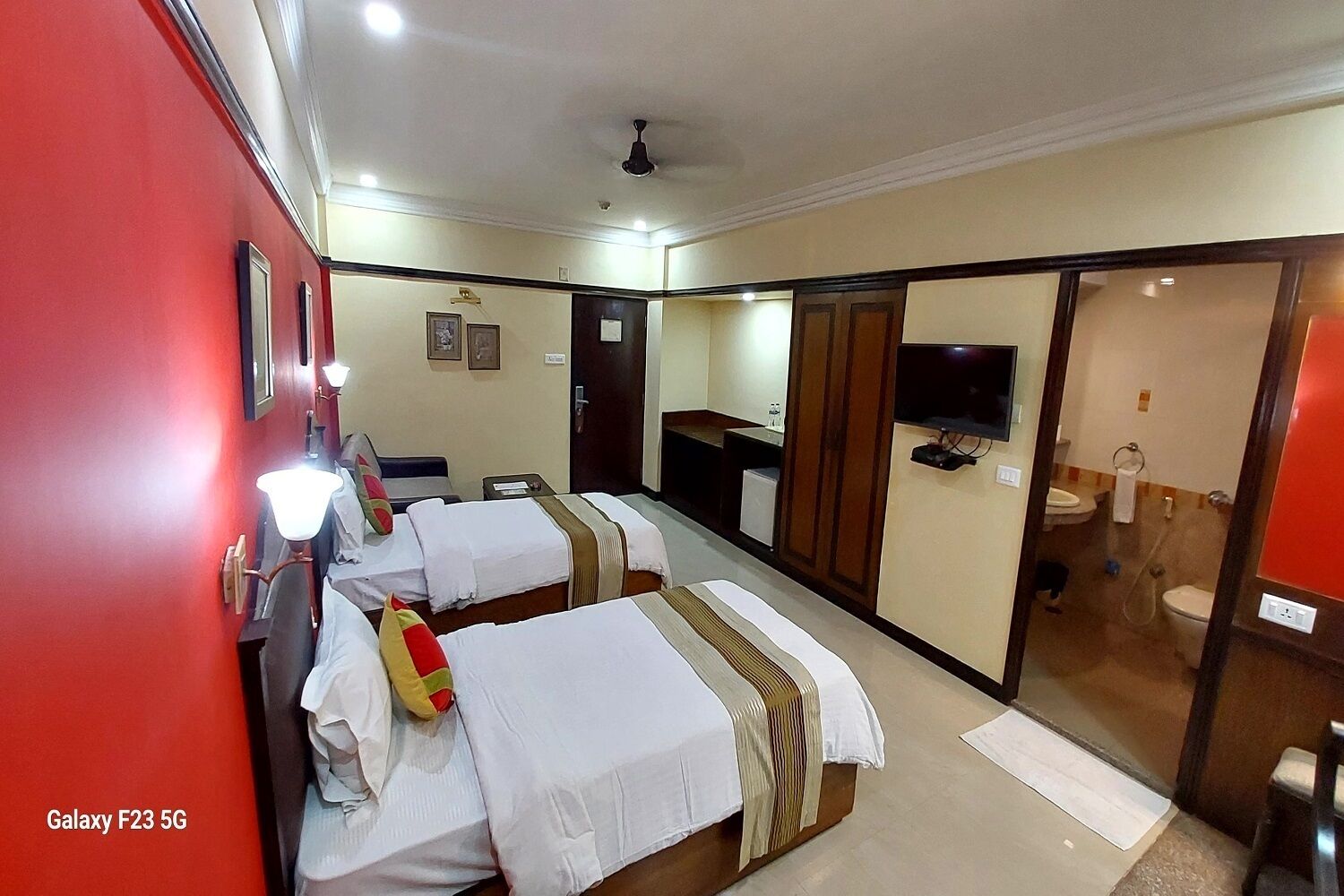 Quarto de hotel com duas camas, televisão, banheiro com vaso sanitário e pia, e uma parede vermelha.