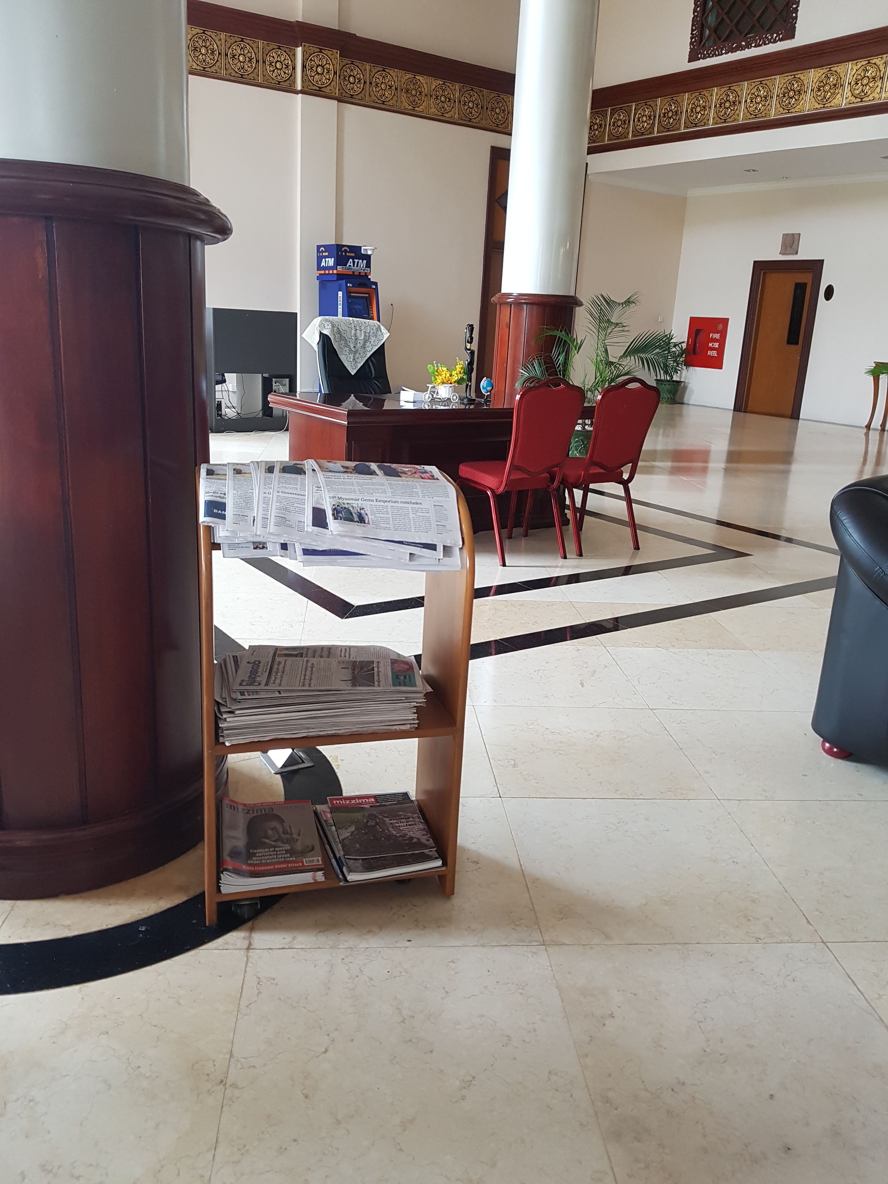 Um hall de entrada com balcão de recepção, cadeiras vermelhas, uma grande coluna de madeira e uma banca de jornais com revistas.