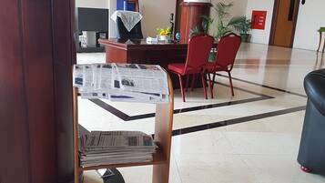 Um hall de entrada com balcão de recepção, cadeiras vermelhas, uma grande coluna de madeira e uma banca de jornais com revistas.