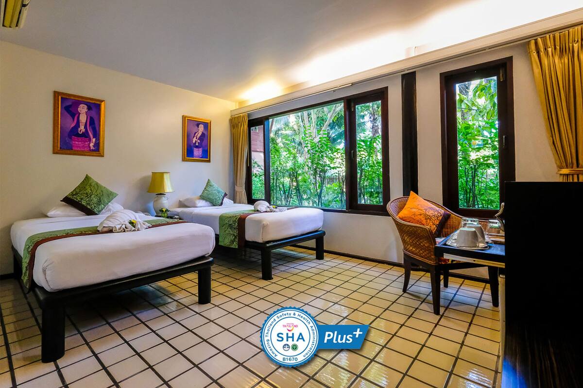 Quarto de hotel com duas camas, uma mesa de jantar e amplas janelas com vista para o verde.