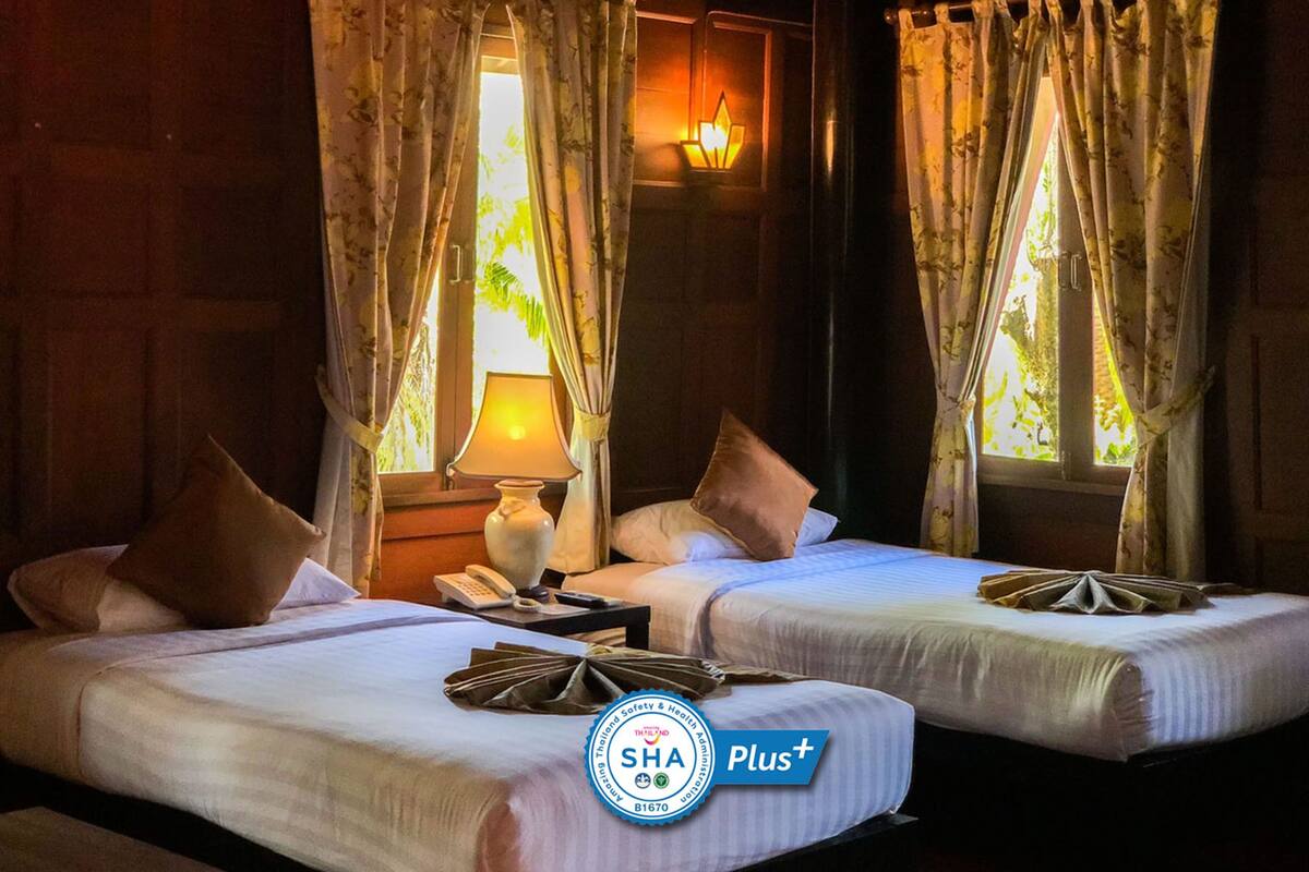 Quarto de hotel com duas camas, uma mesinha de cabeceira, uma luminária e cortinas.