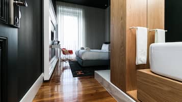 Quarto de hotel moderno com piso de madeira, cama com lençóis brancos, guarda-roupa de madeira e vista da cidade através de amplas janelas.