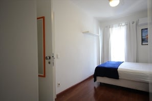 Quarto pequeno e simples, com uma cama, uma porta e uma janela com cortinas.