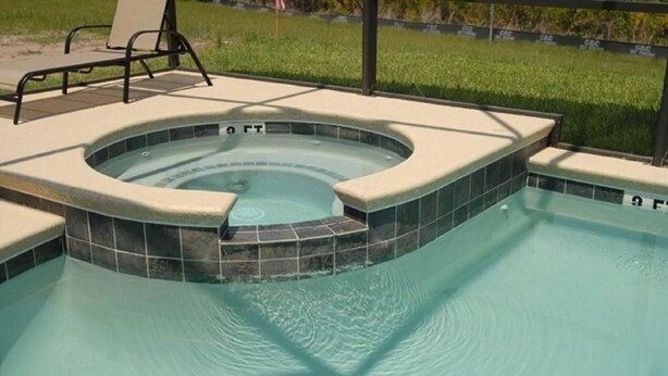 Piscina com área de hidromassagem, rodeada por um deck revestido e uma área gramada ao fundo.