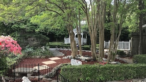 Um jardim com um gazebo, estátuas de pedra e uma área cercada com cobertura de aparas e caminhos de cascalho.