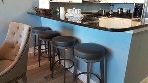 Cozinha com bancada azul, pia branca e torradeira. Há três banquetas pretas e uma cadeira bege.