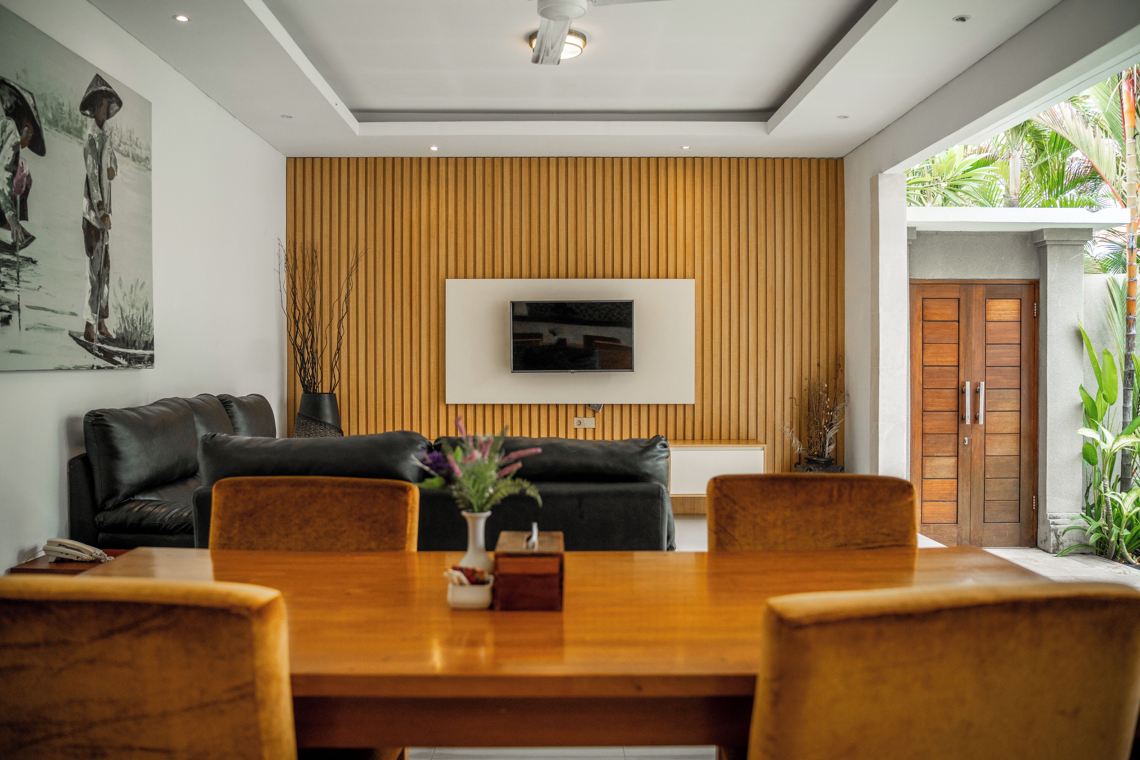Uma sala de estar moderna com parede de painel de madeira, TV montada, mesa de jantar com cadeiras, sofá e mesa de centro com um vaso.