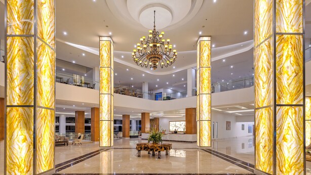Um hall amplo com um lustre central, colunas de mármore e um espaço de estar com uma planta.