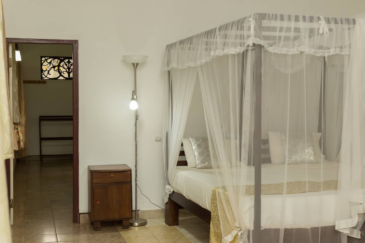 Quarto com cama de dossel, mesa de cabeceira, luminária de chão e um detalhe decorativo na parede.