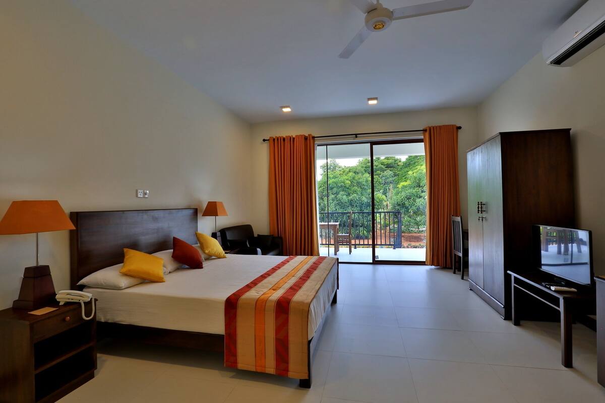 Quarto de hotel com cama, luminárias de cabeceira, escrivaninha com televisão e varanda com cortinas.
