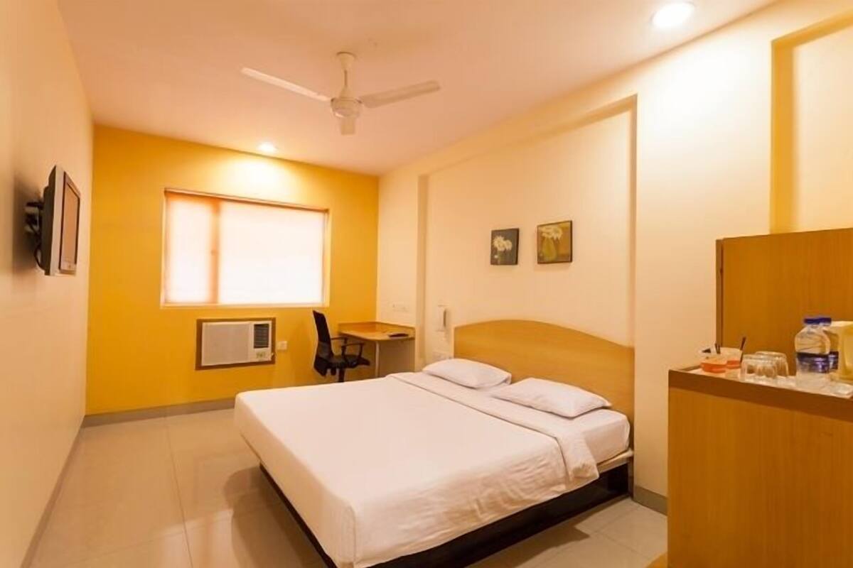 Quarto de hotel com cama, escrivaninha, cadeira, ventoinha no teto e janela com persianas.