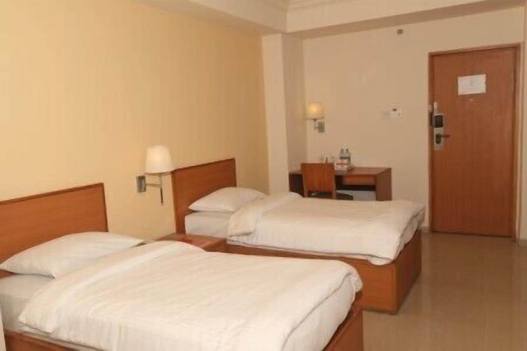 Quarto de hotel com duas camas de solteiro, uma mesa com cadeira, uma luminária e uma porta.