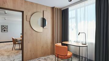 Quarto de hotel moderno com parede de painel de madeira, um espelho redondo, uma pequena mesa redonda com uma lâmpada e vista através de grandes janelas.