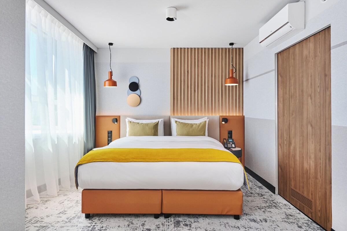 Quarto de hotel moderno com uma cama grande, cabeceira de madeira, mesa de cabeceira com telefone e uma janela ampla com cortinas.