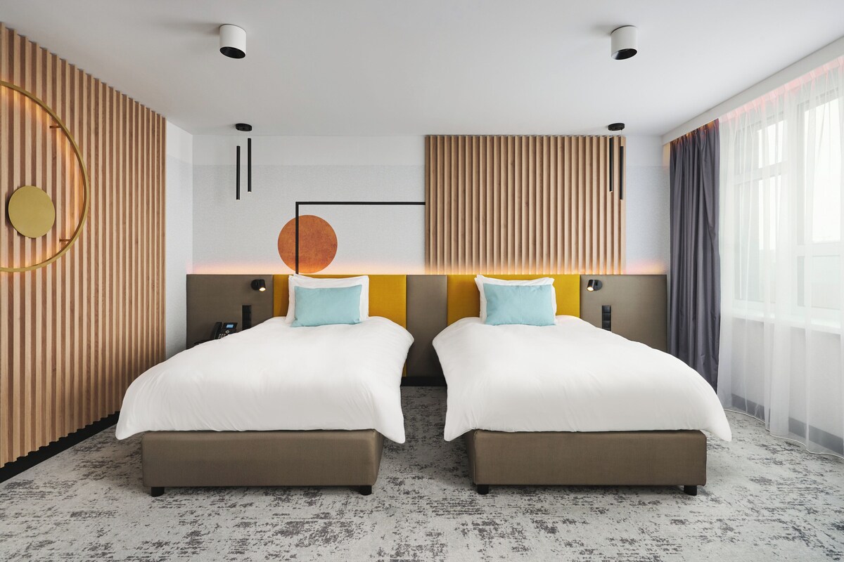 Quarto de hotel moderno com duas camas, parede de destaque em madeira, uma janela grande com cortinas e um luminário circular.