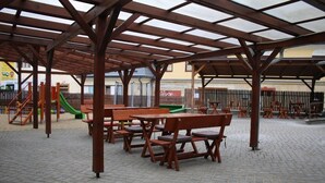 Uma área de varanda coberta ao ar livre com bancos e mesas de madeira, um parquinho infantil com escorrega e piso de tijolos.
