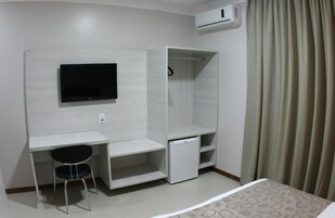 Quarto de hotel compacto com cama, escrivaninha com cadeira, televisão, guarda-roupa e pequeno frigorífico.