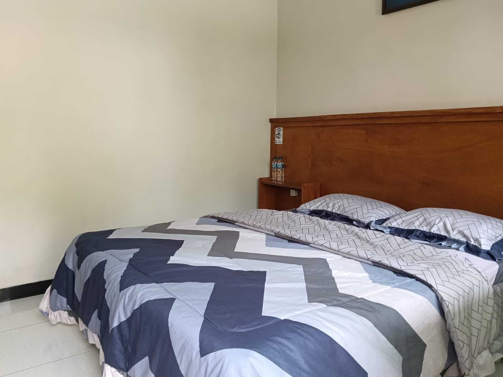 Uma cama bem arrumada com um cobre-leito estampado, uma cabeceira de madeira e uma pequena mesa de cabeceira com uma garrafa de água.