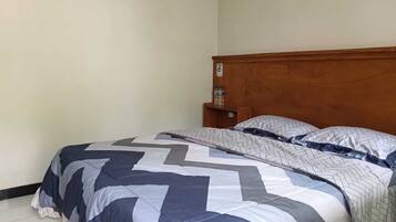Uma cama bem arrumada com um cobre-leito estampado, uma cabeceira de madeira e uma pequena mesa de cabeceira com uma garrafa de água.