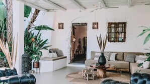 Uma sala de estar moderna com sofá, mesa de centro e cadeiras. Há plantas e uma janela com grades.