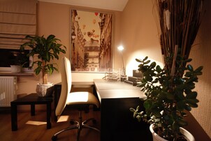 Quarto com escrivaninha, cadeira, plantas em vasos e uma grande moldura na parede.