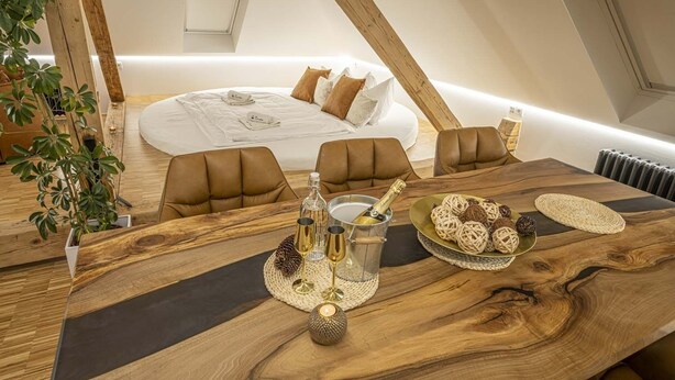 Quarto estilo loft aconchegante, com uma cama grande, uma mesa de jantar posta com champanhe, copos e objetos decorativos.