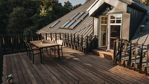Um deck de madeira com mesa e cadeiras, uma cerca de metal e um edifício com janelas amplas e telhado inclinado.
