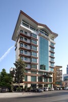Um hotel de vários andares com uma placa grande 'MASEL HOTEL', uma área de estacionamento com vários carros e um céu claro ao fundo.