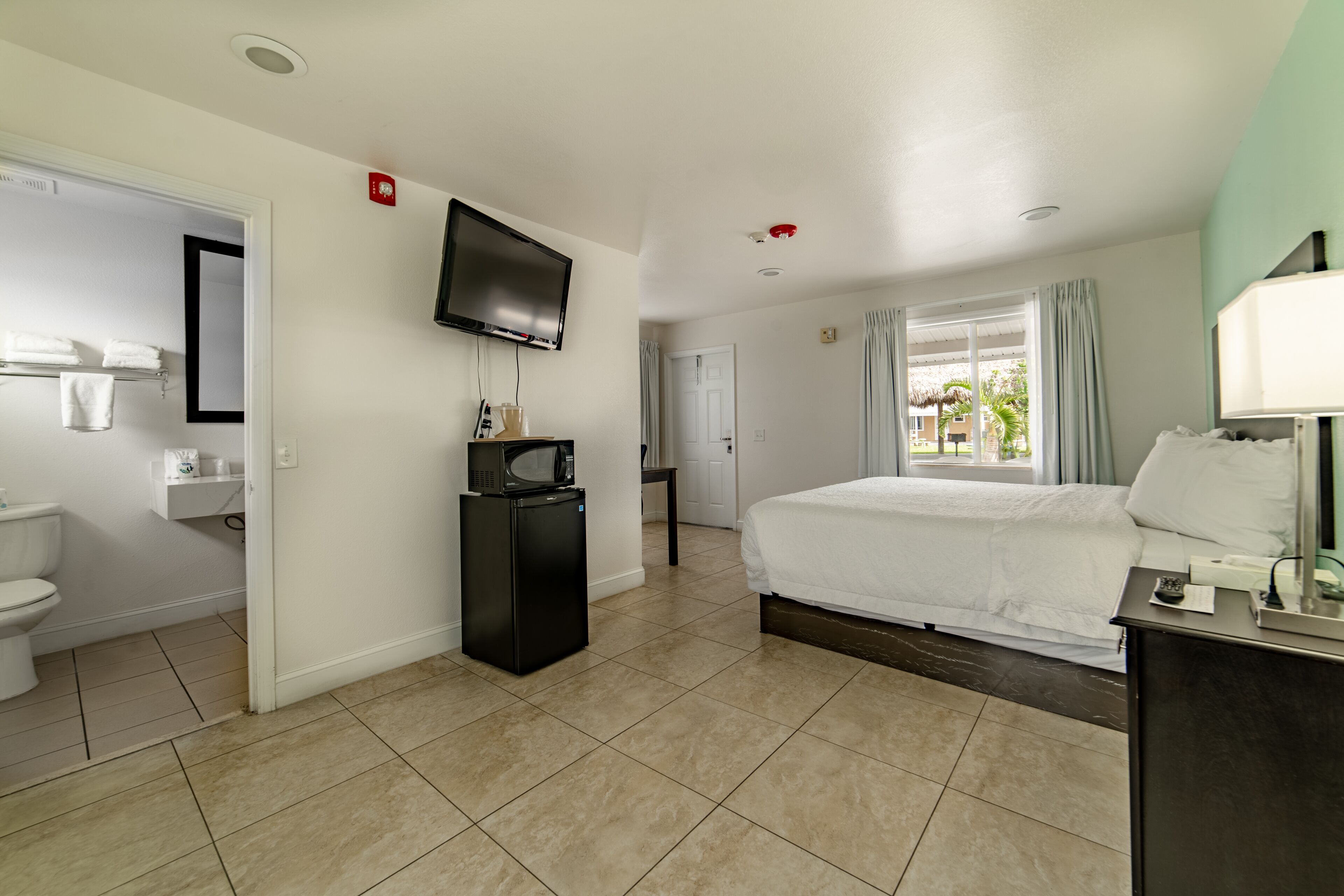 Quarto de hotel com cama, televisão na parede, pequena cozinha e banheiro com vaso sanitário e pia.