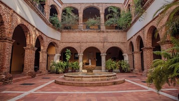Pátio com uma fonte central, rodeado por arcos de tijolo e vasos de plantas.