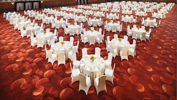 Um amplo salão de banquetes com mesas redondas e cadeiras brancas, piso coberto por carpete e cortinas vermelhas.