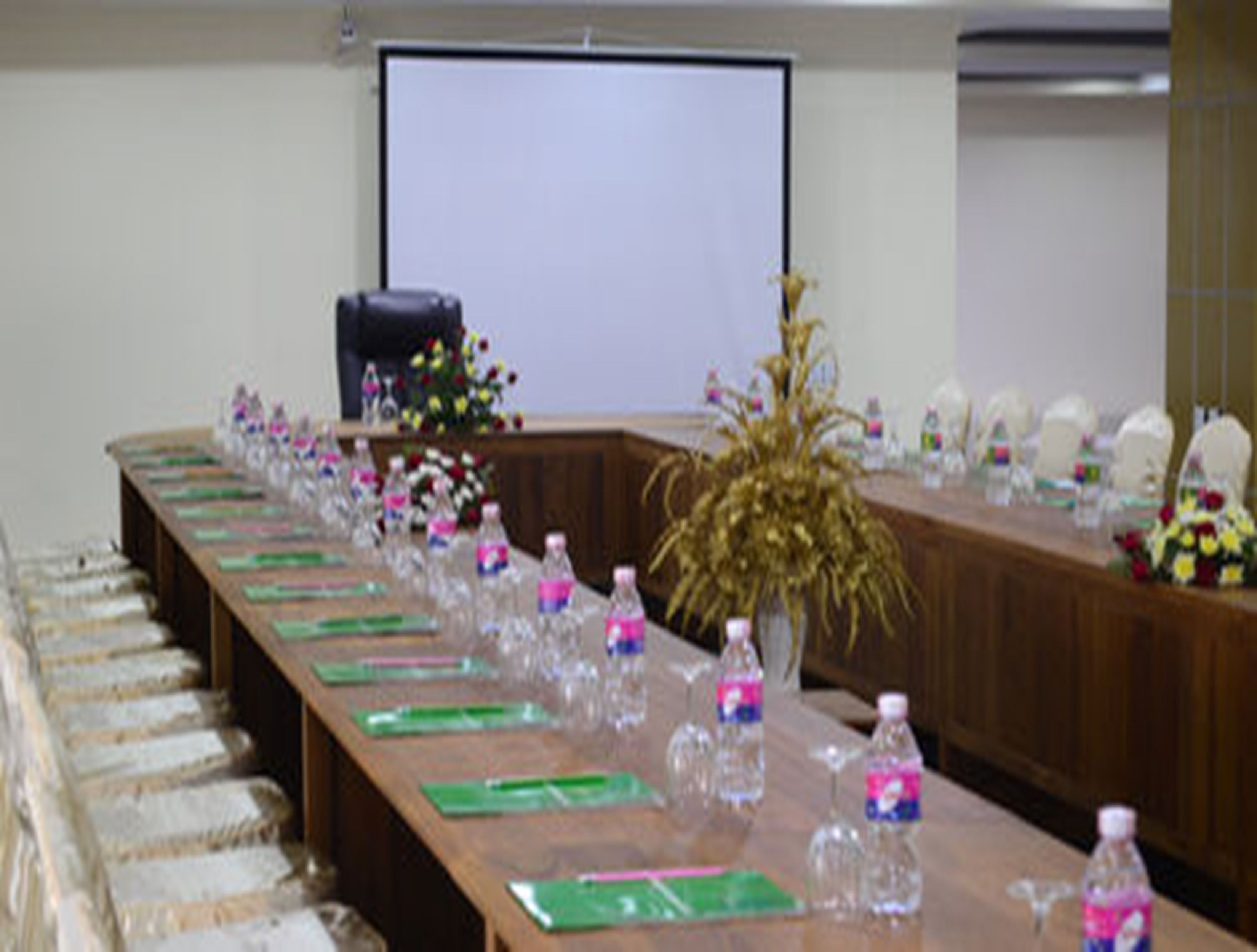 Sala de conferências com uma mesa comprida disposta com garrafas de água e pastas verdes, uma tela de projetor e uma planta decorativa.