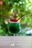 Coquetel em taça com guarnição de frutas vermelhas e uma fatia, sobre fundo verde desfocado.