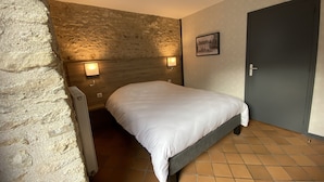 Quarto com parede de pedra, cama com roupa de cama branca, duas luzes fixadas na parede e uma porta.