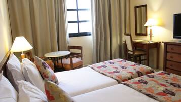 Quarto de hotel com duas camas, uma escrivaninha, uma cadeira, uma mesinha e uma televisão.