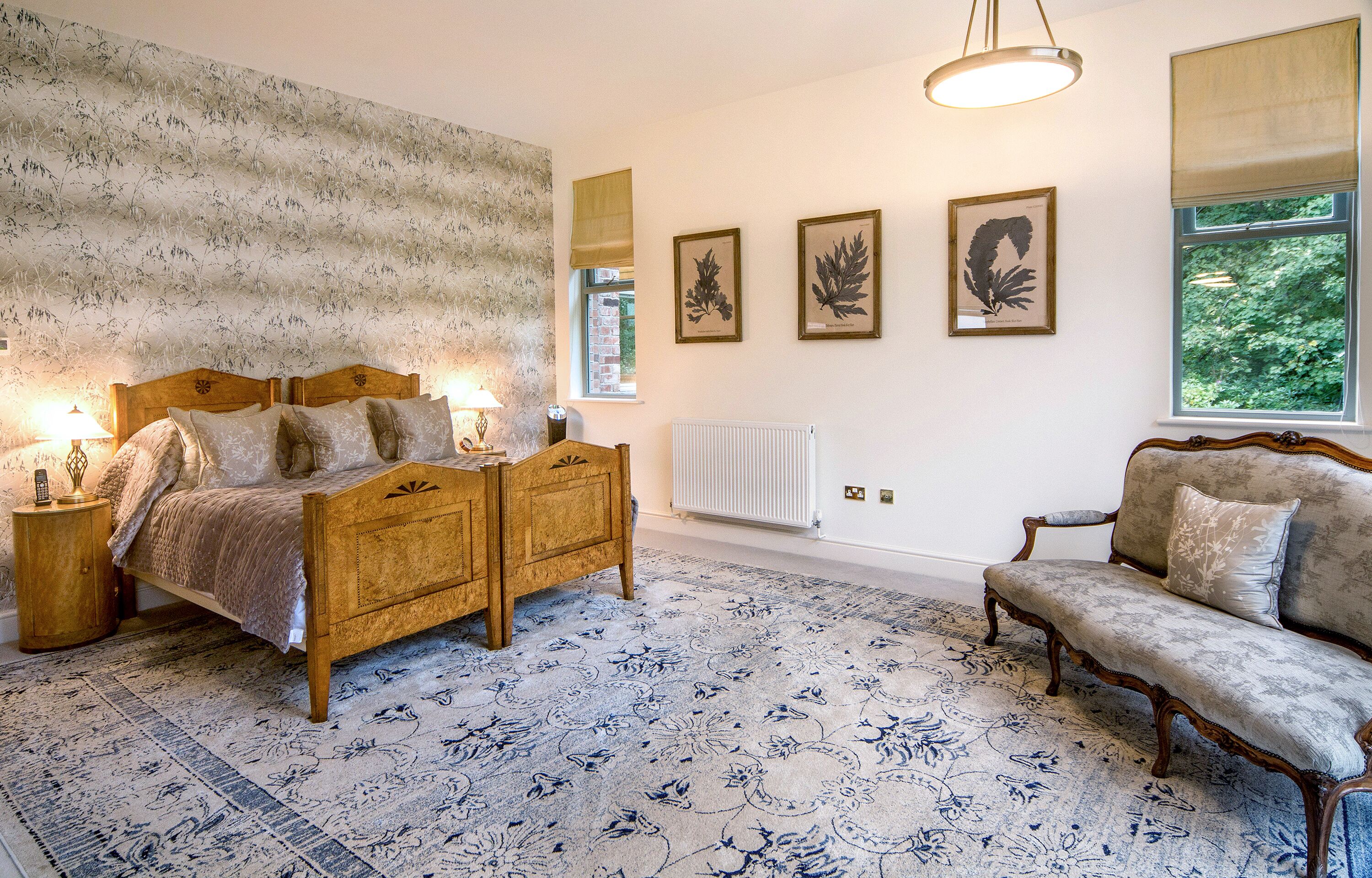 Quarto com cama de madeira, um sofá, tapete estampado e gravuras botânicas emolduradas na parede.